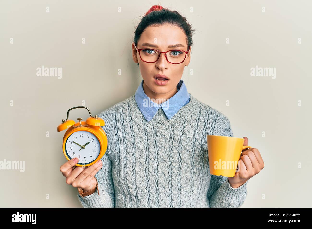 Young caucasian woman holding alarm clock drinking coffee clueless and ...