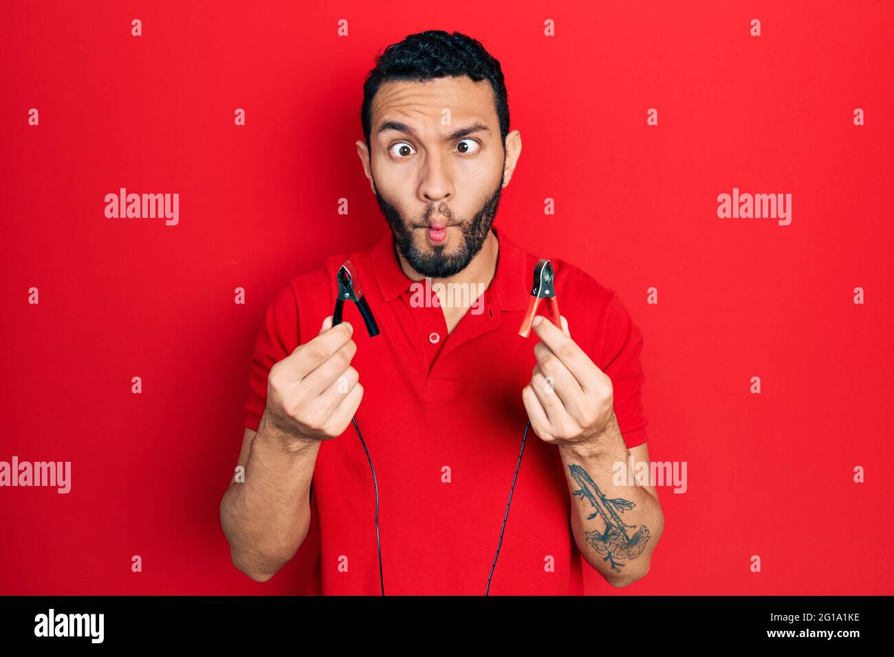Hispanic man with beard holding battery clamps making fish face with ...