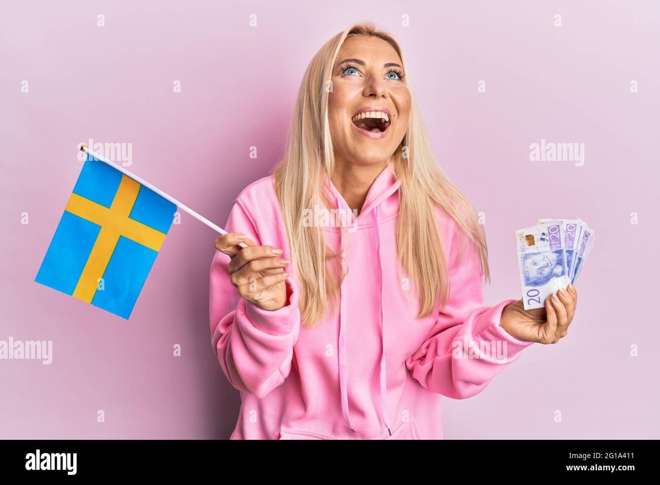Young blonde woman holding sweden flag and krone banknotes angry and ...