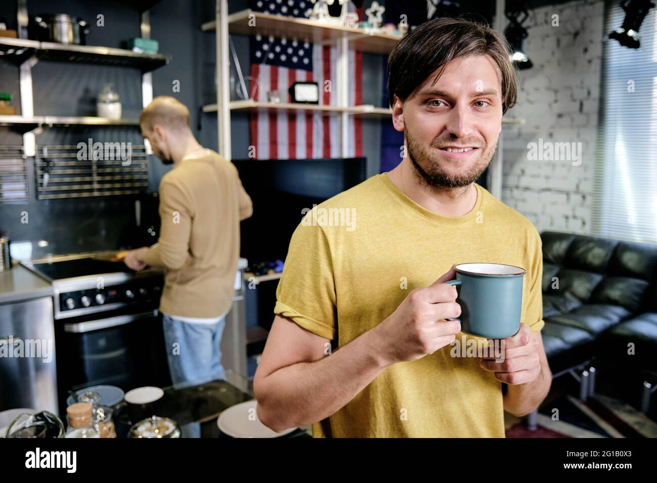 Smiling guy having drink against man cooking breakfast in the kitchen ...