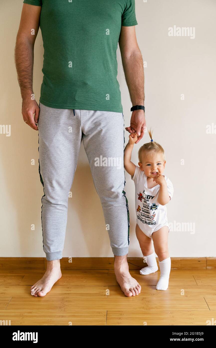 Dad holds the hand of a little toddler with a ponytail standing on the ...