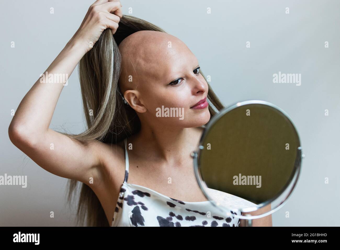 Young confident lady with alopecia looking in mirror and taking off wig ...