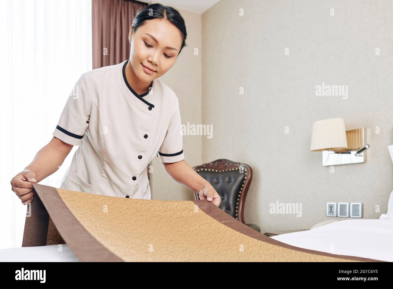Pretty smiling hotel maid making bed and putting on bed runner as a ...