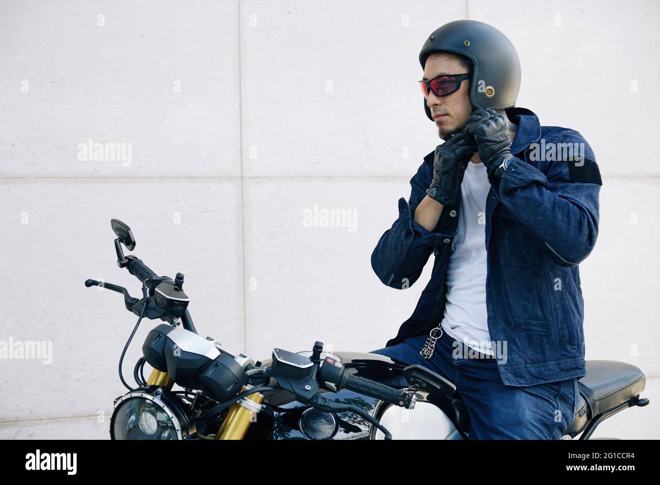 Motorcyclist in denim jacket fastening helmet before riding on highway ...