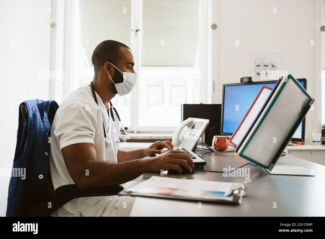 Male doctor using computer while sitting at desk during COVID-19 Stock ...