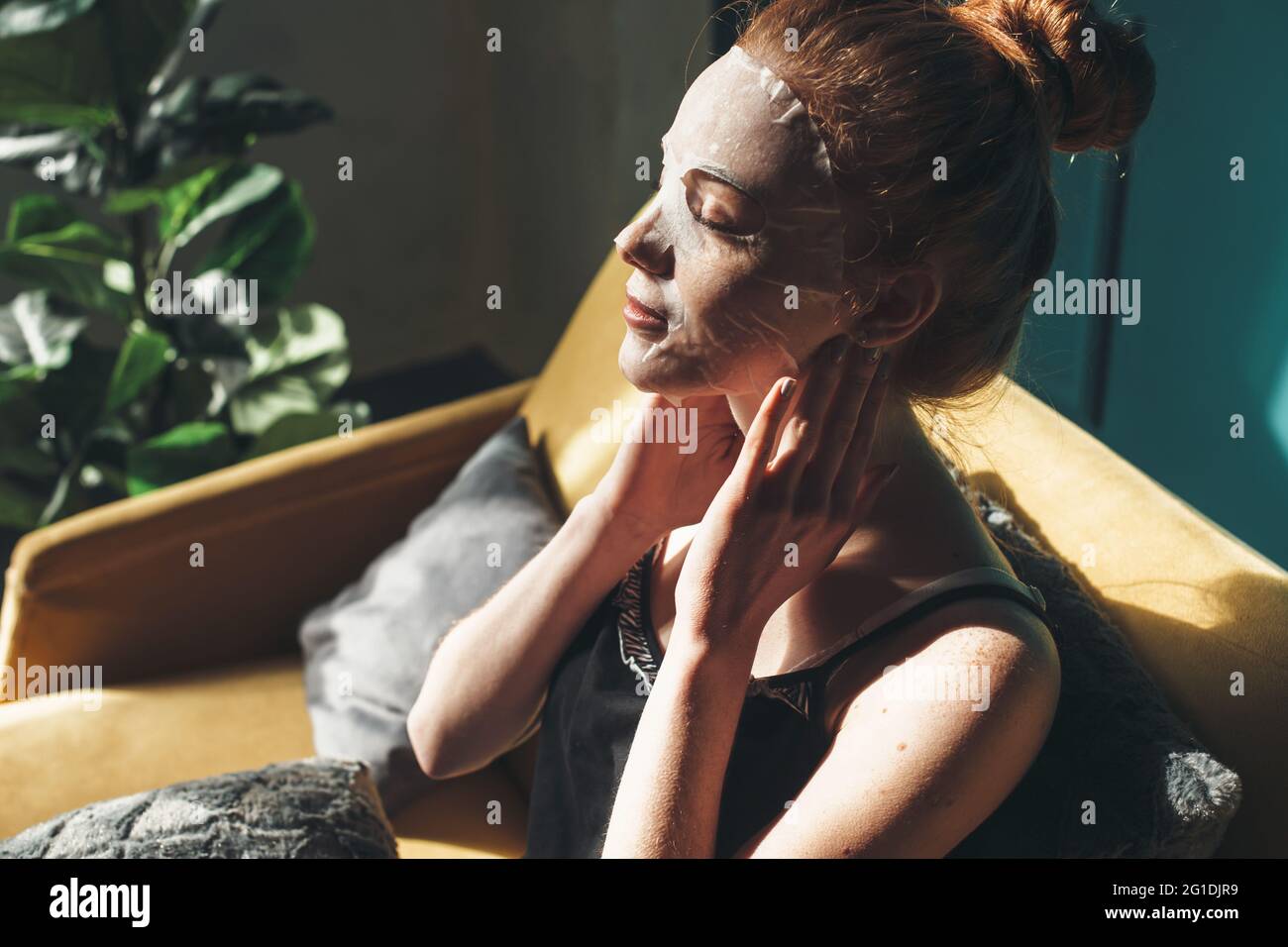 Ginger woman is applying a paper mask on facial skin during home spa ...