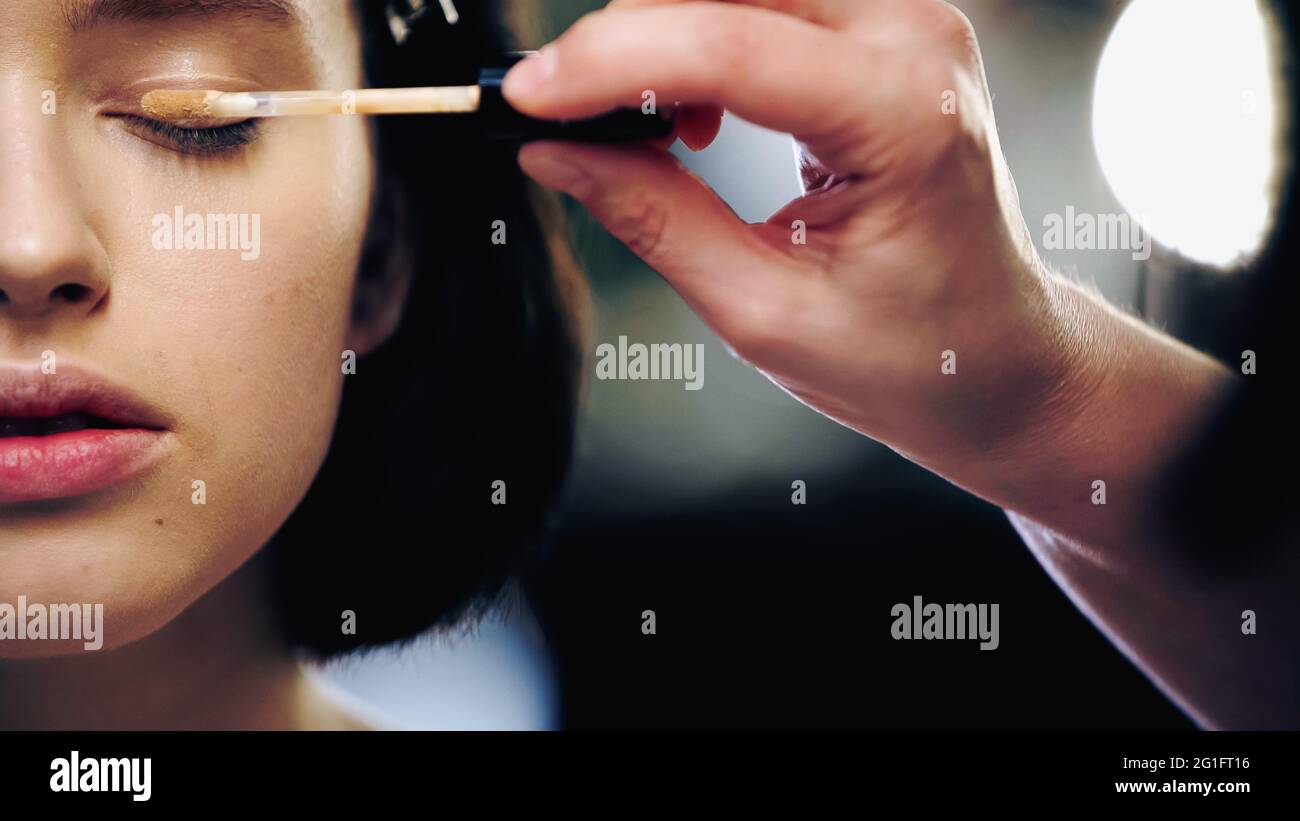 cropped view of makeup artist applying concealer on eyelid of model ...