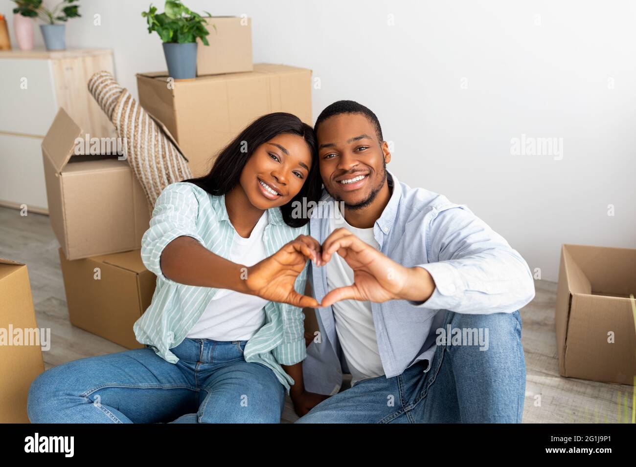 Love nest. Happy black couple sitting among moving boxes, making heart ...