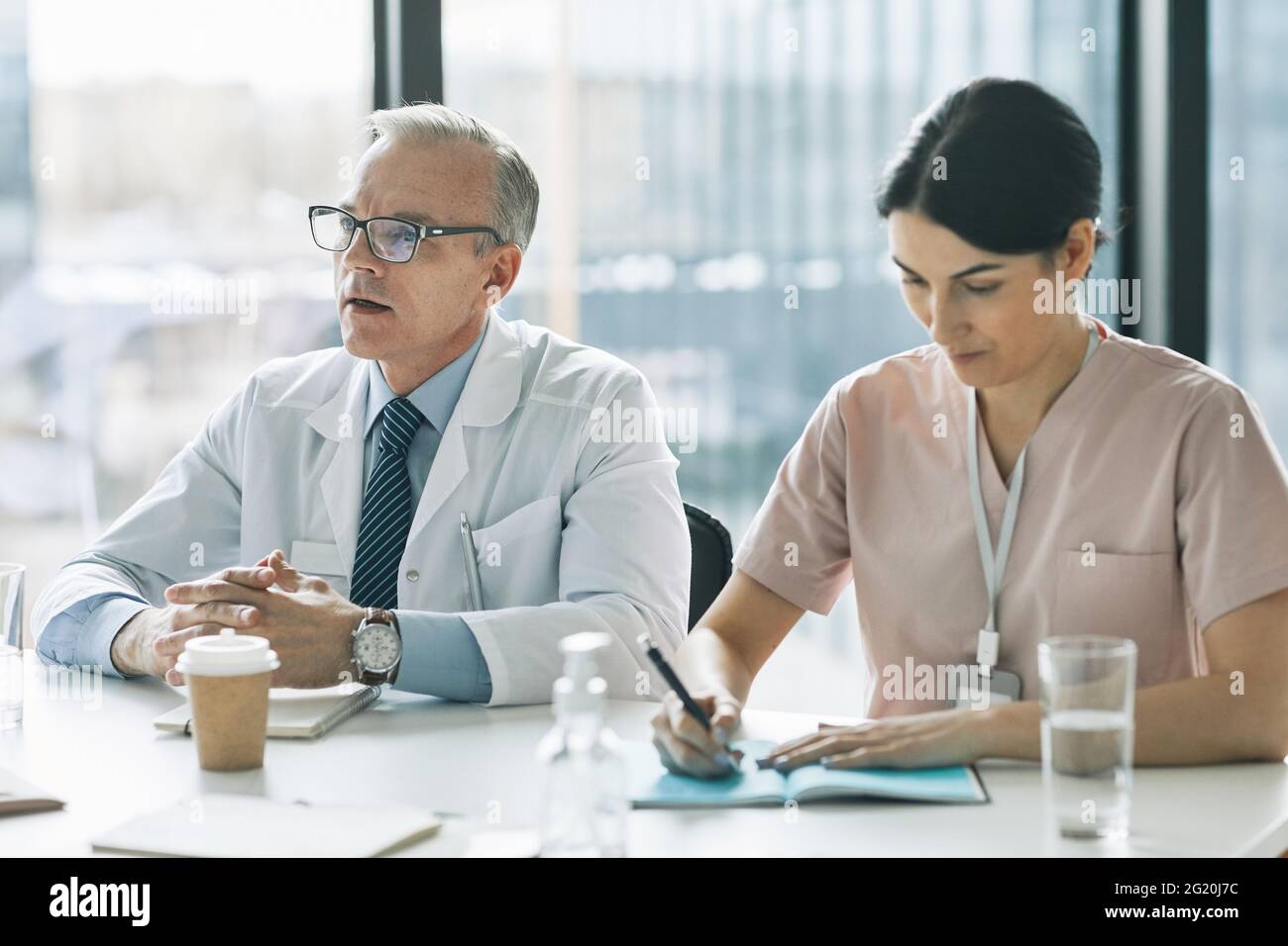 Portrait of two doctors sitting at meeting table by window and taking ...