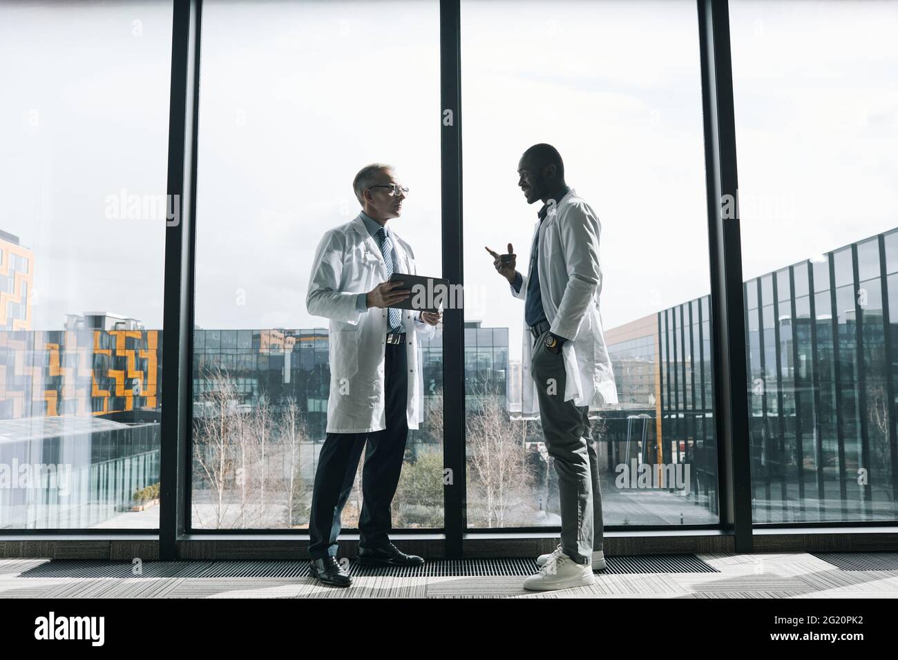 Full length portrait of two doctors talking while standing by window in ...