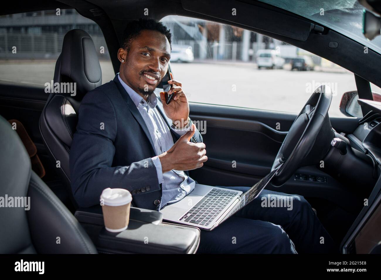 Smiling african man in business suit sitting in modern car with laptop ...