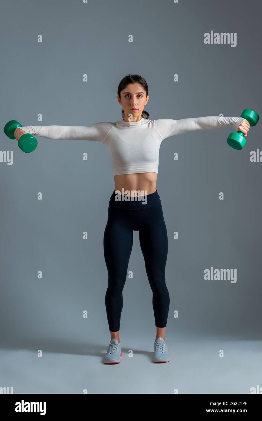 Athletic girl performing dumbbell lateral raise on grey background ...