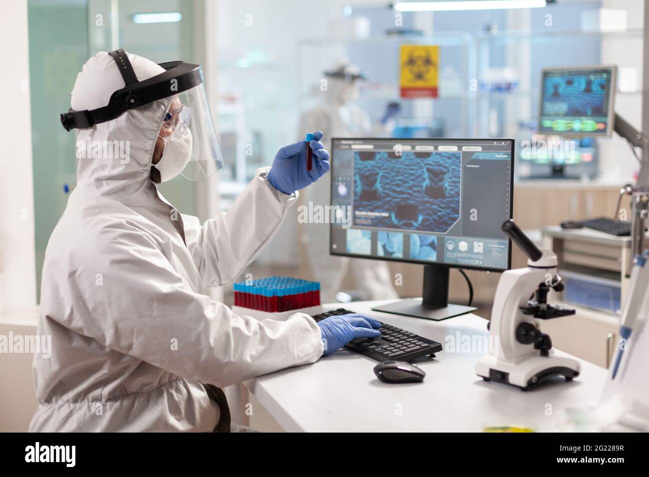 Scientist dressed in ppe suit working on computer holding vaccutainer ...