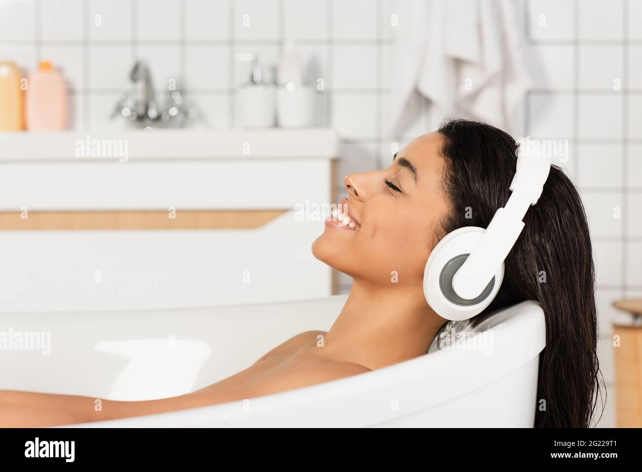 happy young woman lying in bathtub with closed eyes and listening music ...
