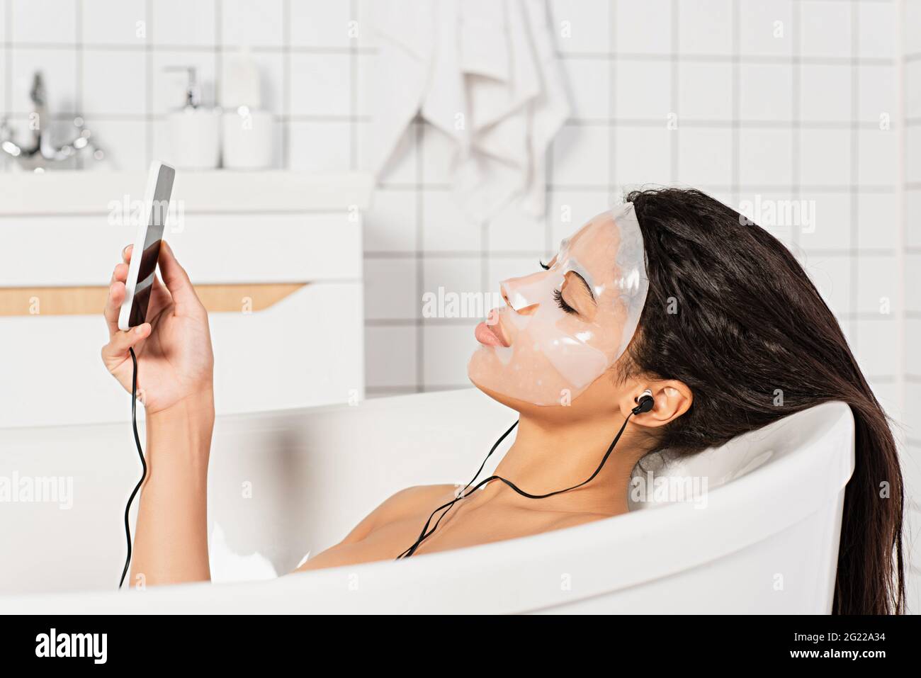 relaxed young woman with sheet mask on face lying in bathtub with ...