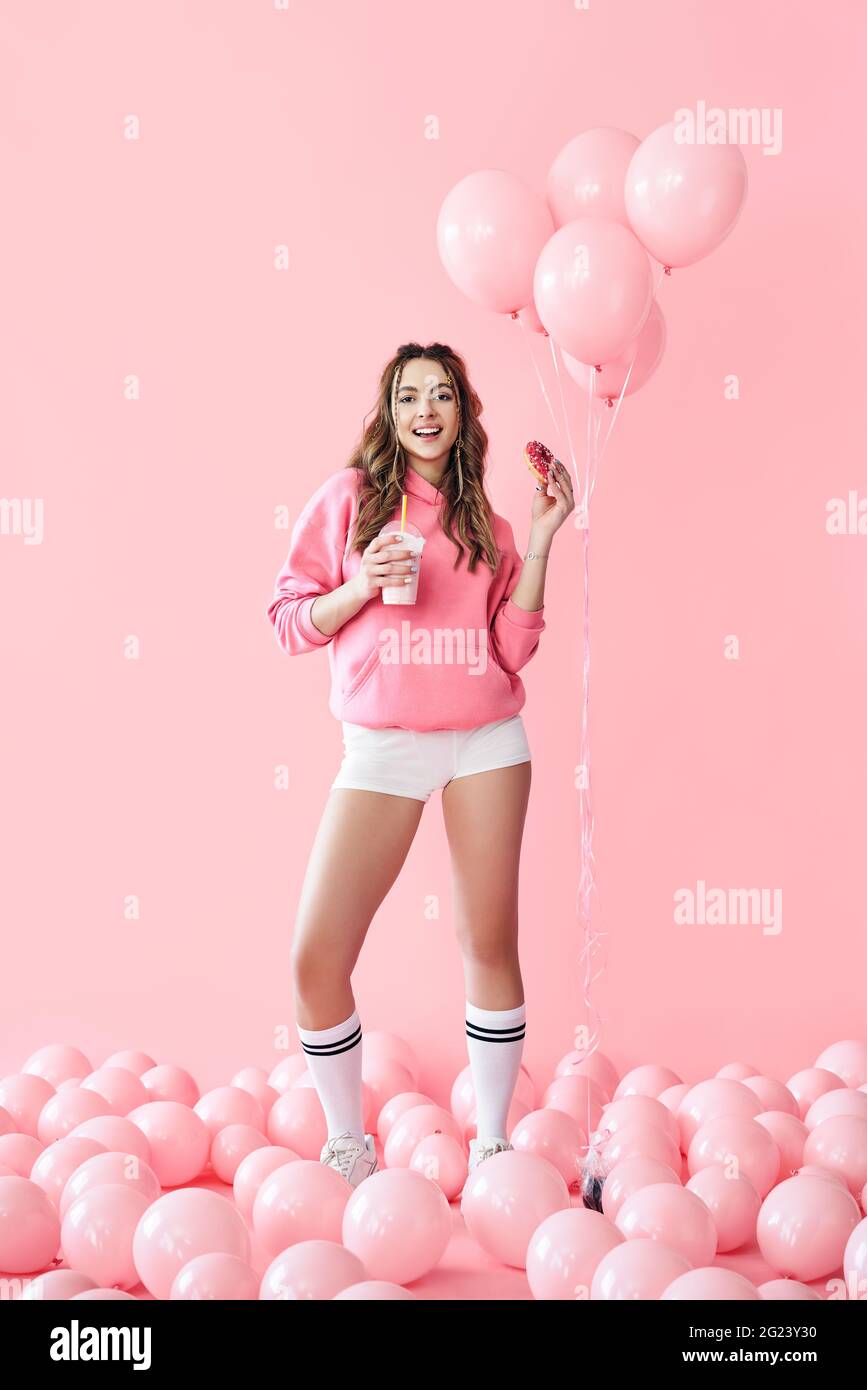 Happy young woman with milkshake and donut in hands standing over many ...
