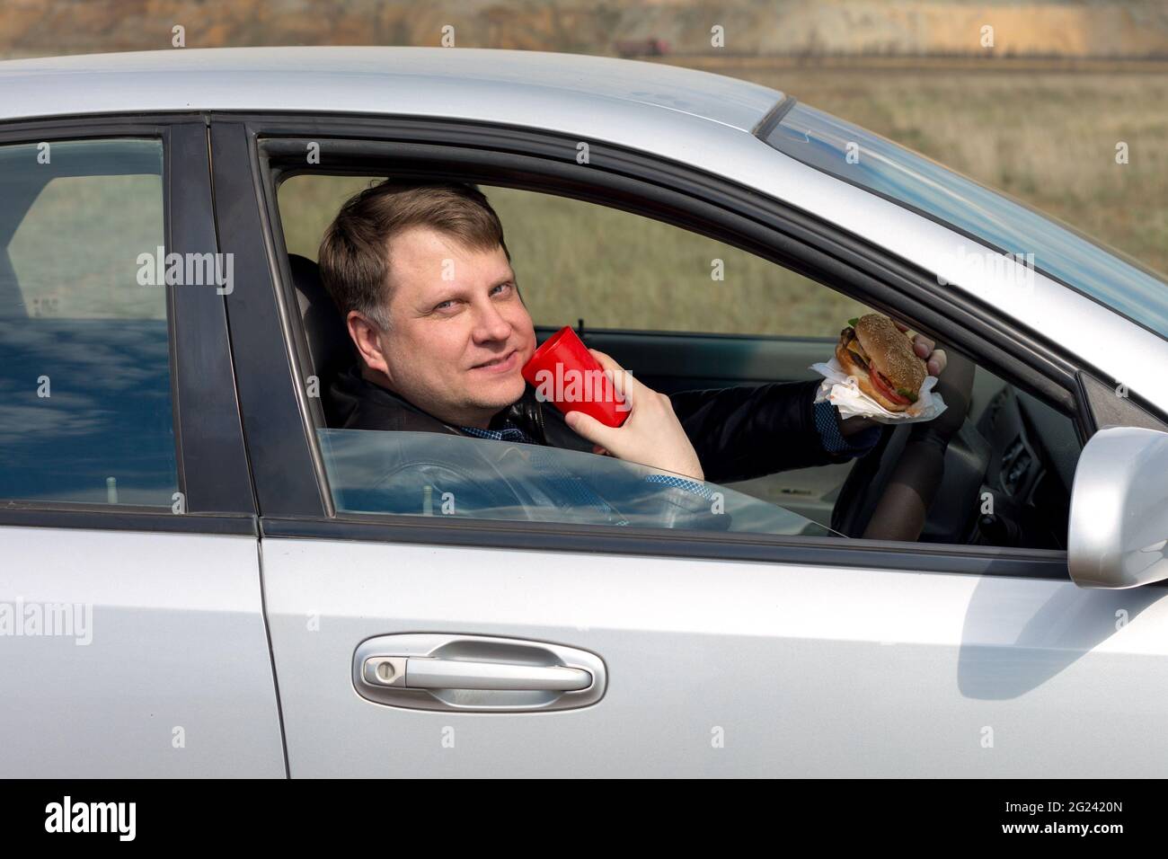 Happy man driving a car with a glass and hamburger in his hand, goes to ...
