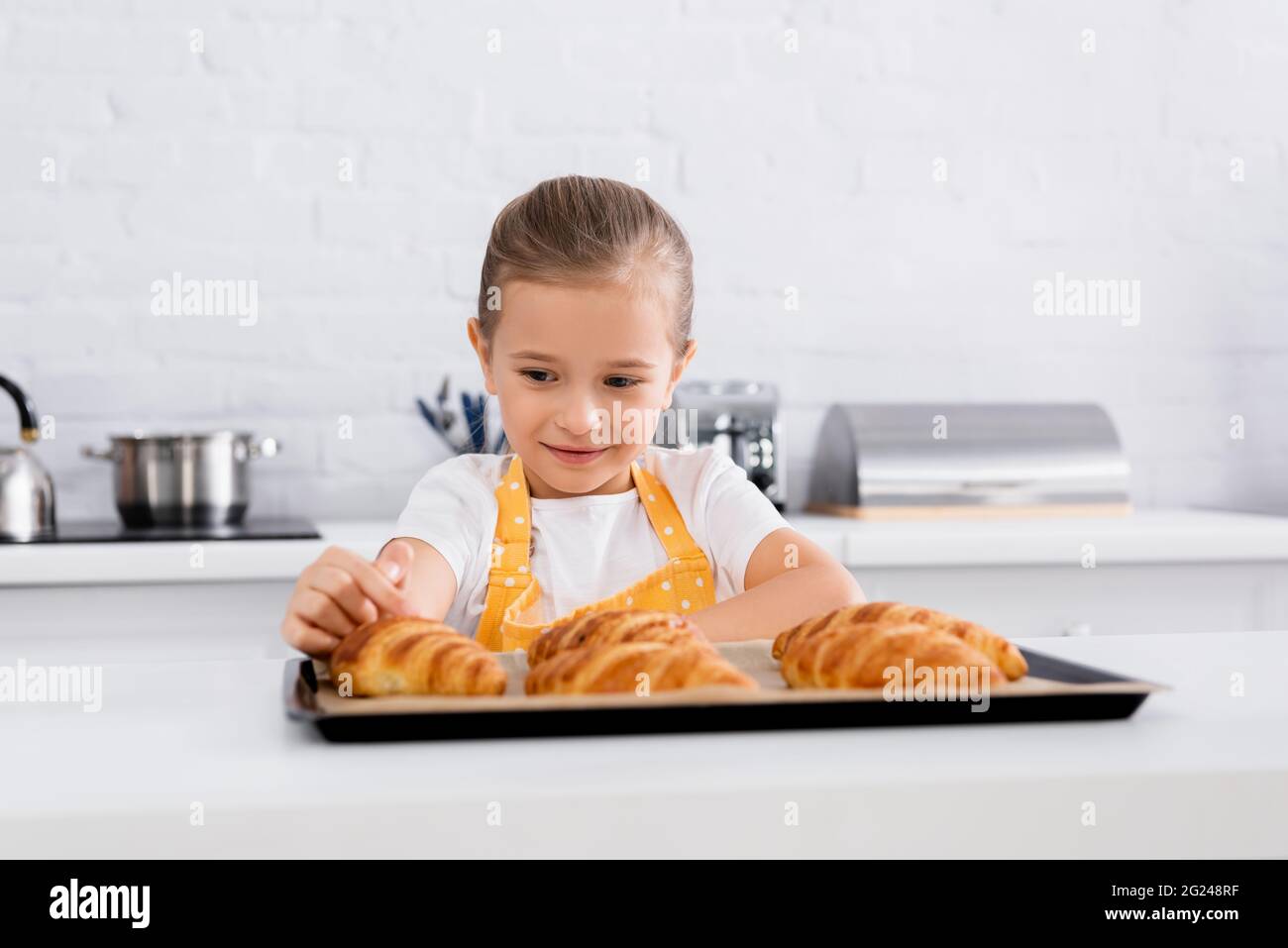 Smiling kid in apron touching homemade croissant on blurred foreground ...