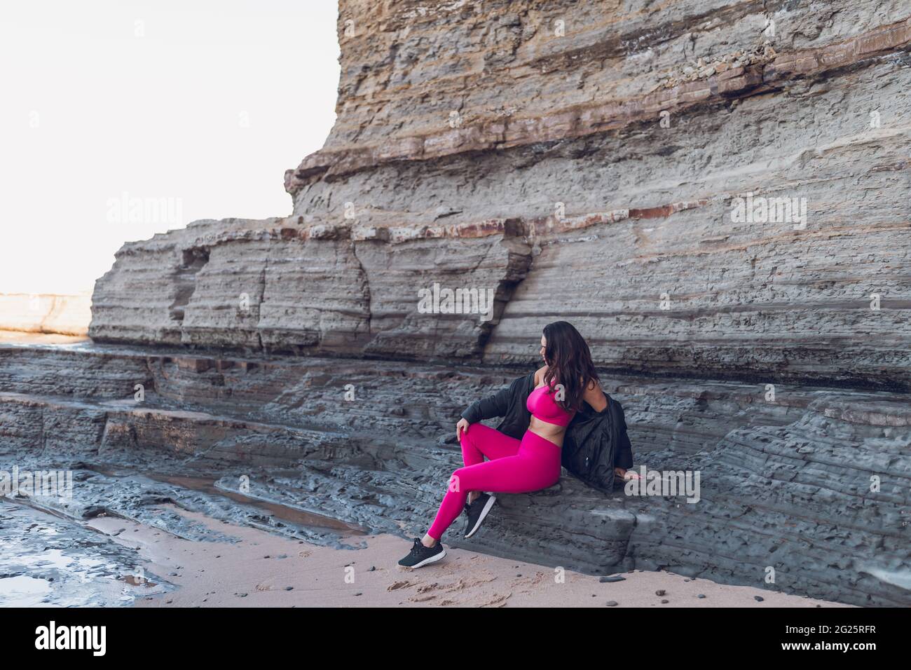 Woman wearing hot pink gym clothes sitting on the rocks at the beach ...