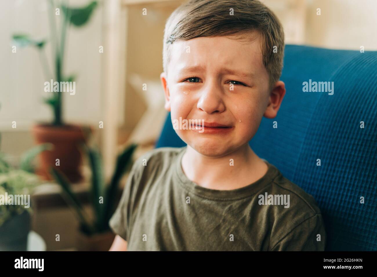 Close up portrait of crying little kid boy, sitting on couch indoors at ...