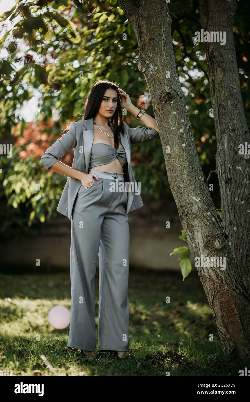 Adult woman wearing a gray suit with a crop top and leaning on a tree ...