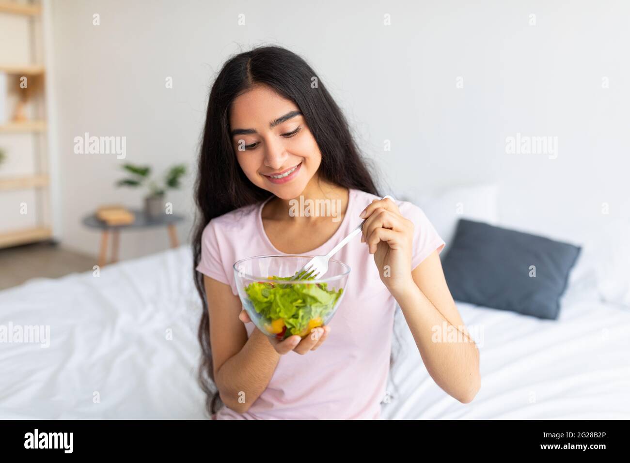 Healthy vegan diet. Joyful Indian lady sitting on bed at home with bowl ...
