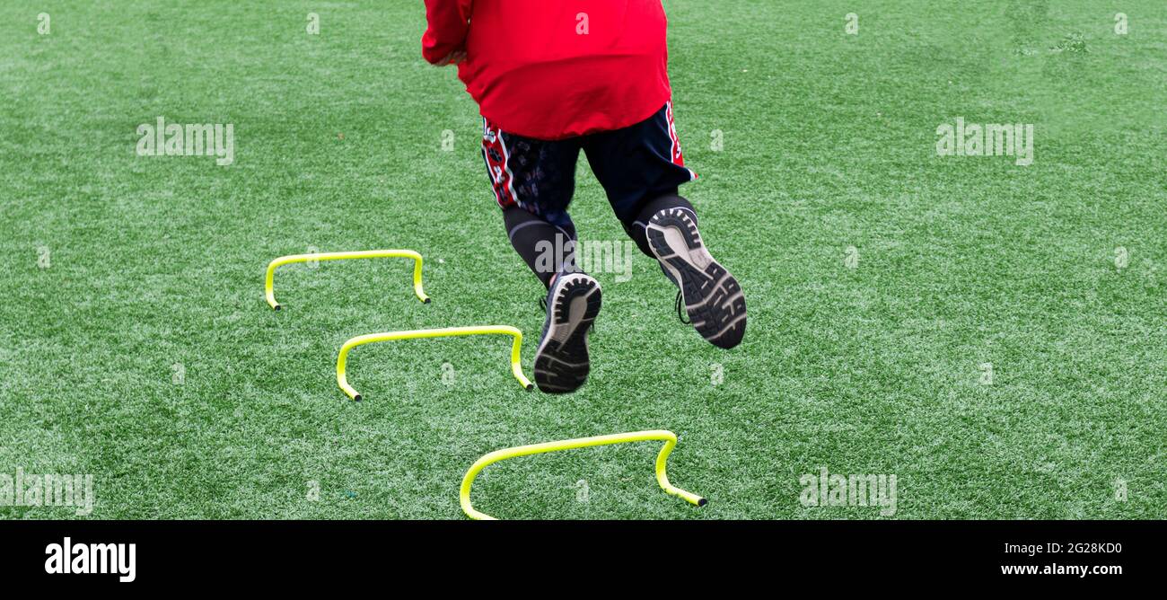 A high school track athlete is jumping over six inch yellow mini hurdles on a green turf field ...