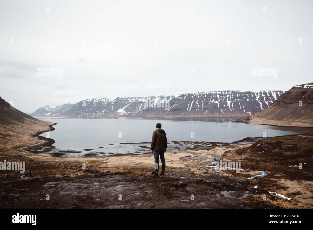 Back view of man standing on shore of lake with cliffs on background ...