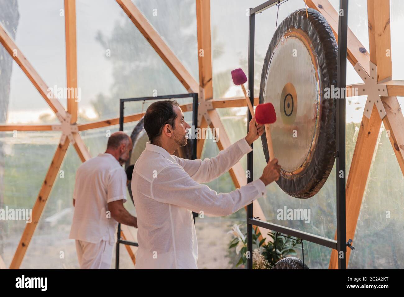Side view of men in white clothes hitting gong in light room with ...