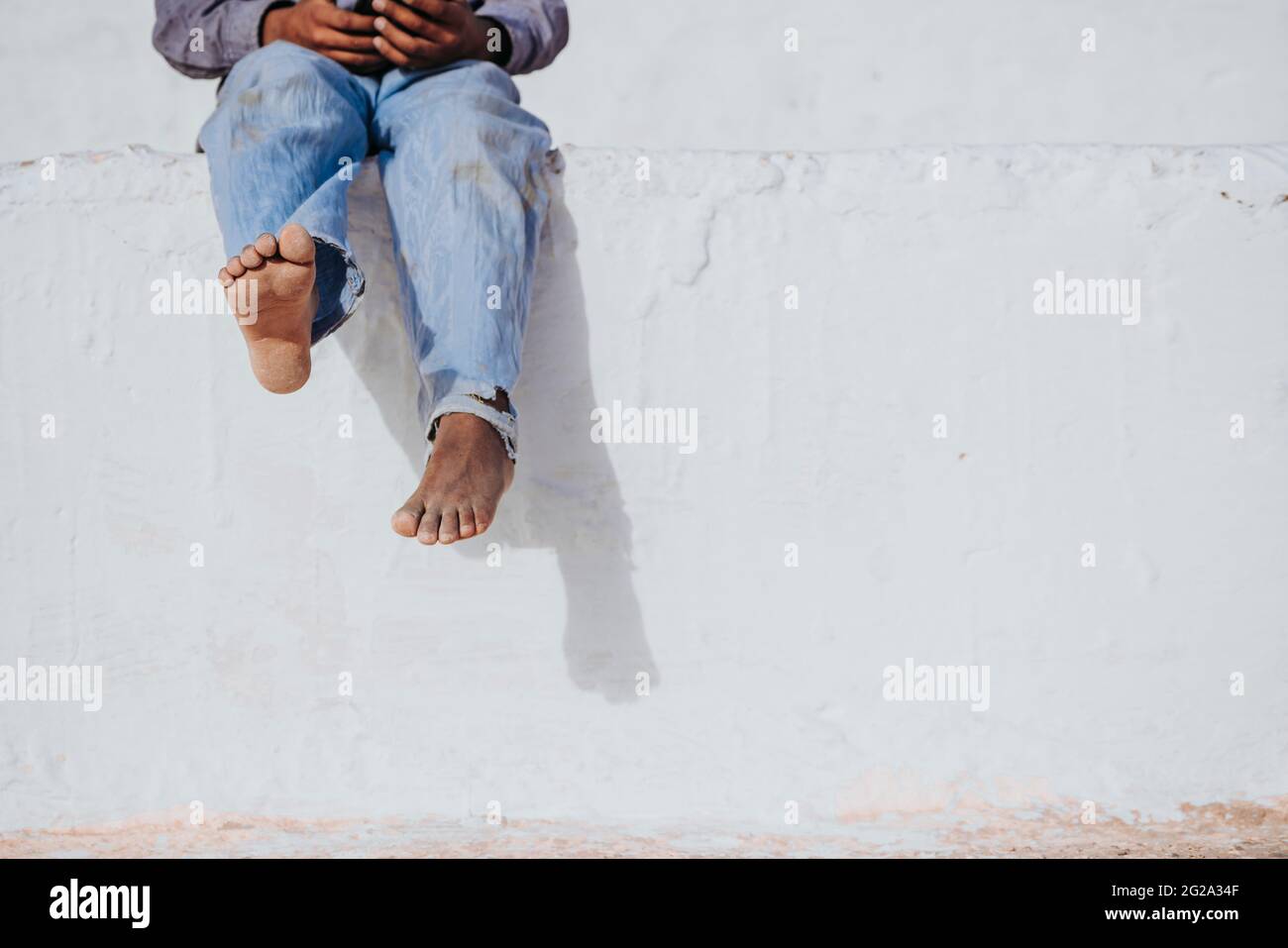 Barefoot legs of anonymous African kid in jeans sitting on shabby white ...