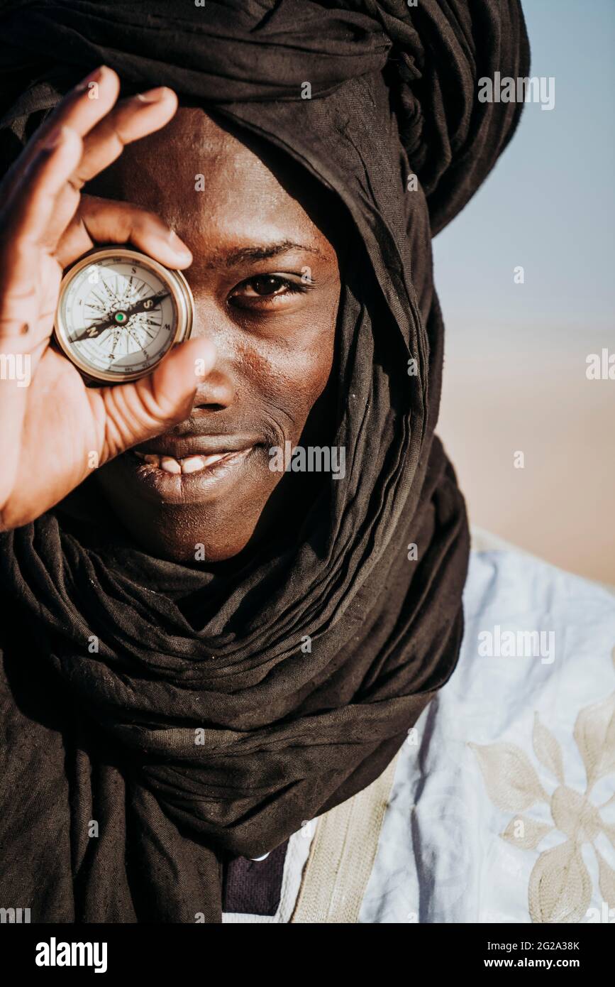 African male in keffiyeh smiling and holding vintage compass near eye ...