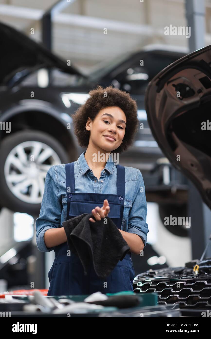 smiling young african american mechanic in overalls drying hands with ...