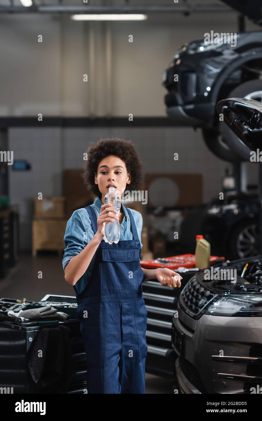 young african american mechanic drinking water from bottle near car in ...