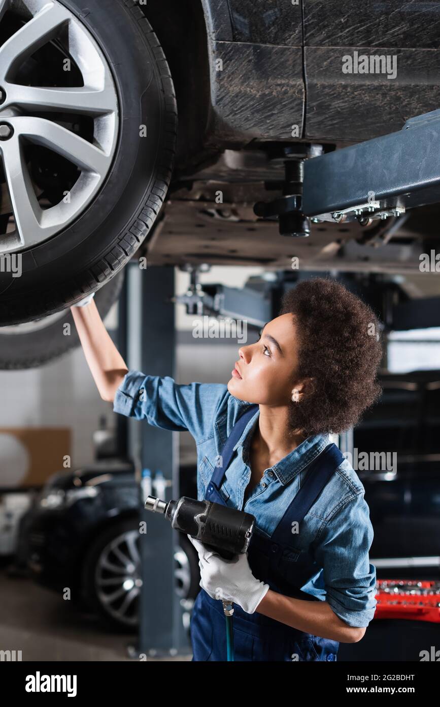 young african american mechanic in overalls repairing wheel on lifted ...