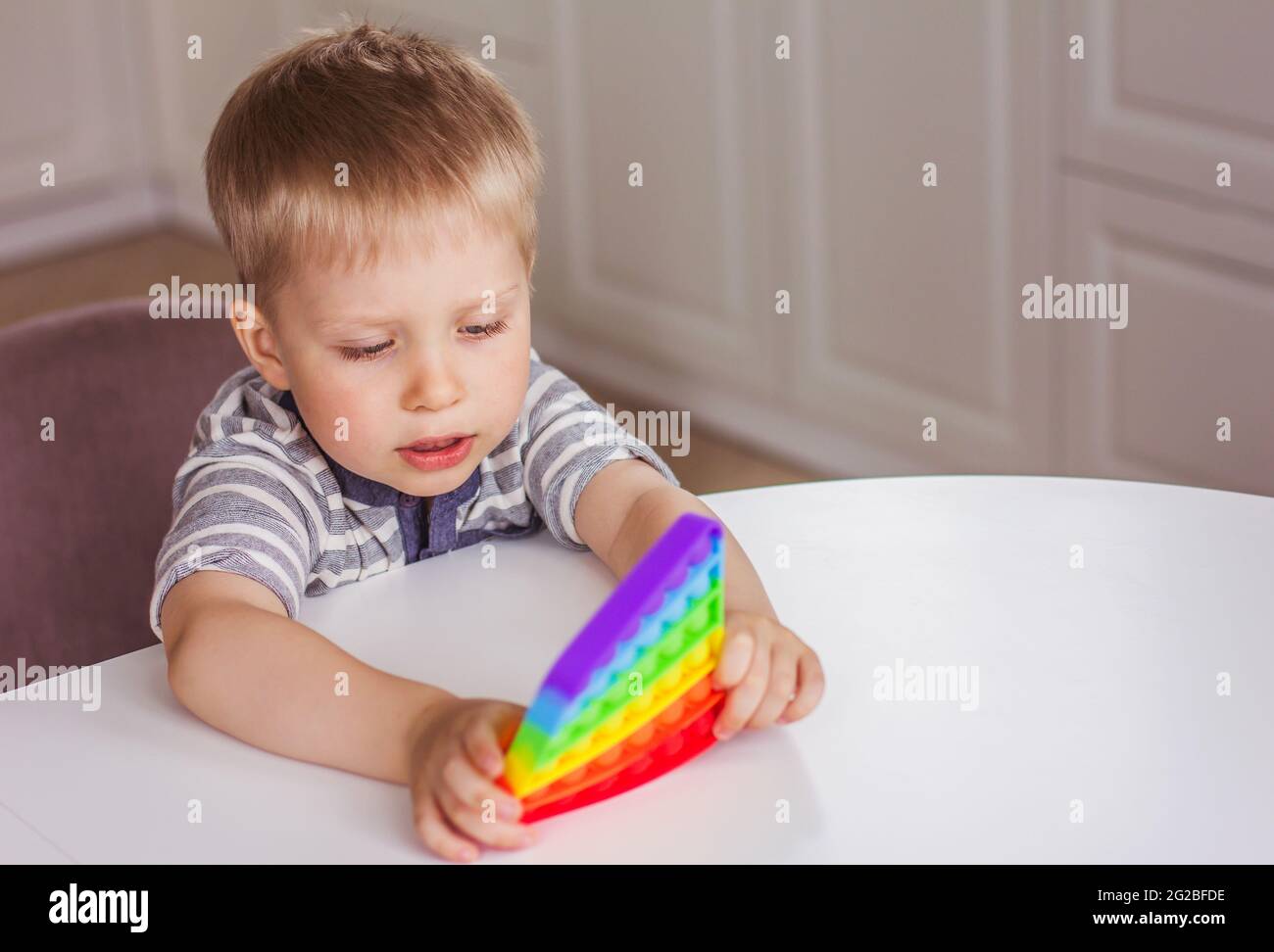Blonde boy playing with rainbow pop it fidget toy. Push bubble fidget ...