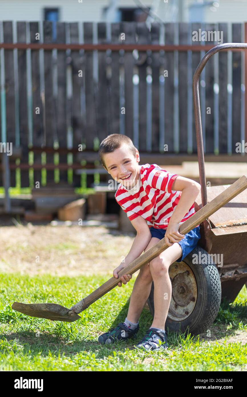 A little cheerful boy sits on a garden wheelbarrow and holds a shovel ...