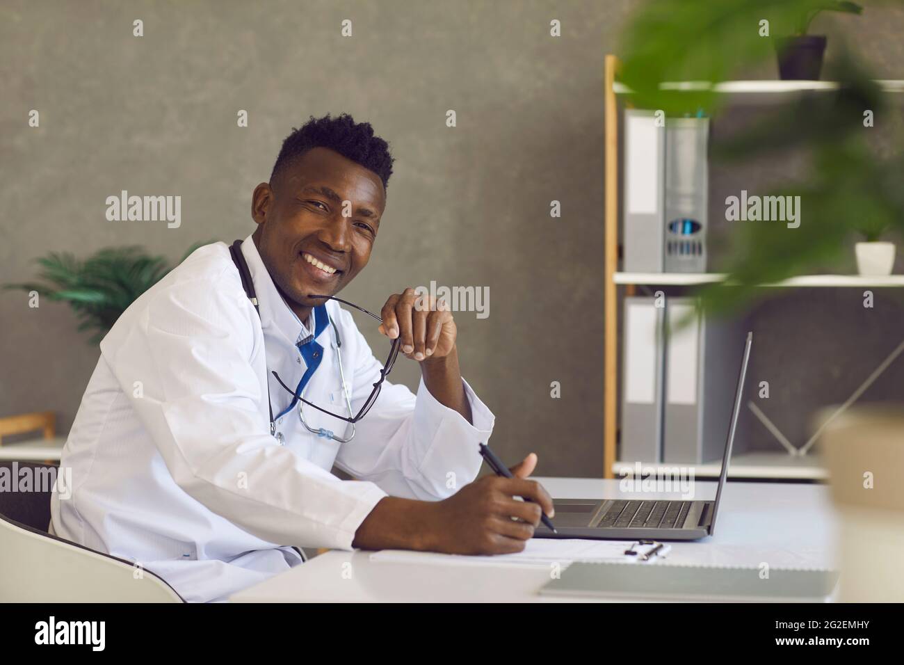 Young black doctor sitting at office desk with laptop, looking at ...