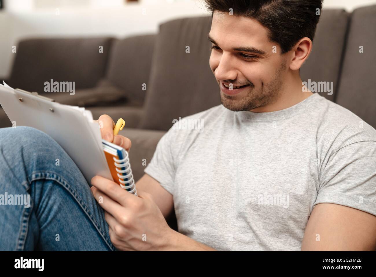 Happy brunette guy writing down notes while sitting on floor at home ...