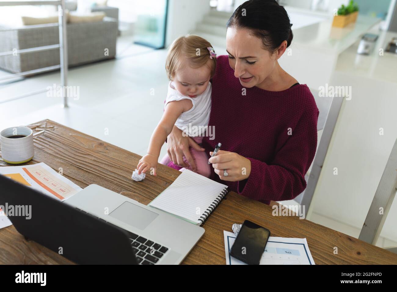 Caucasian mother holding her baby taking notes while working from home ...