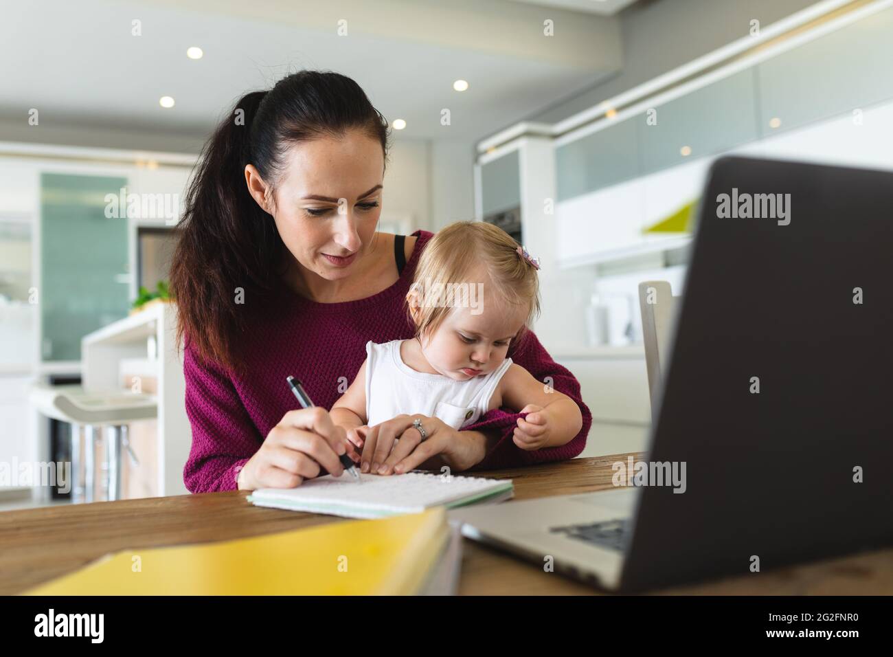 Caucasian mother holding her baby taking notes while working from home ...