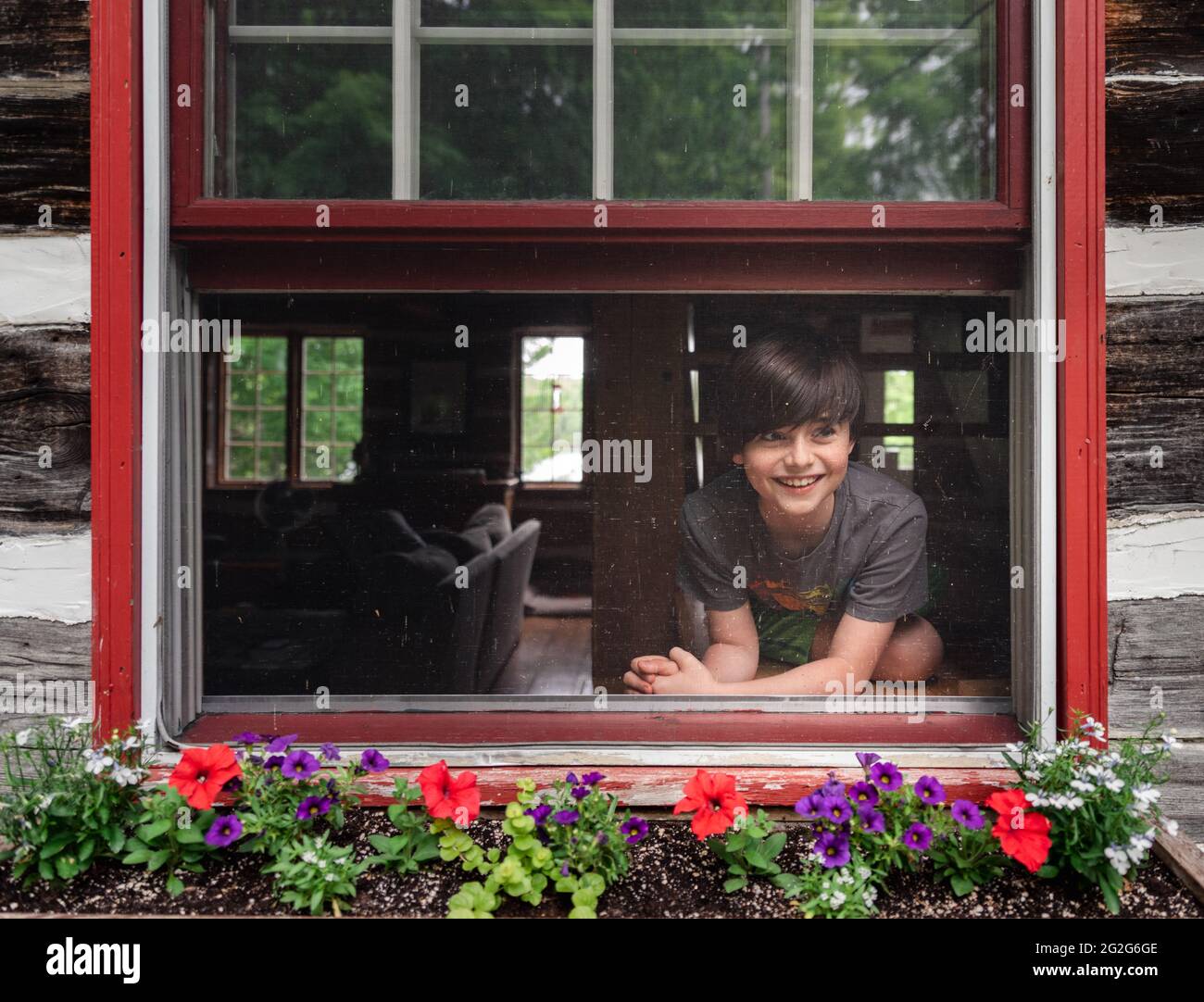 Young happy boy looking out open window of log cabin on summer day ...