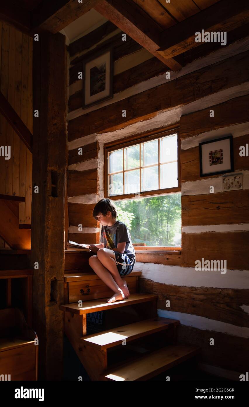 Boy reading a book indoors on the sunny step of a log cabin home Stock ...