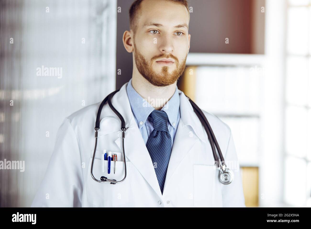 Friendly bearded doctor standing straight in sunny clinic. Portrait of ...