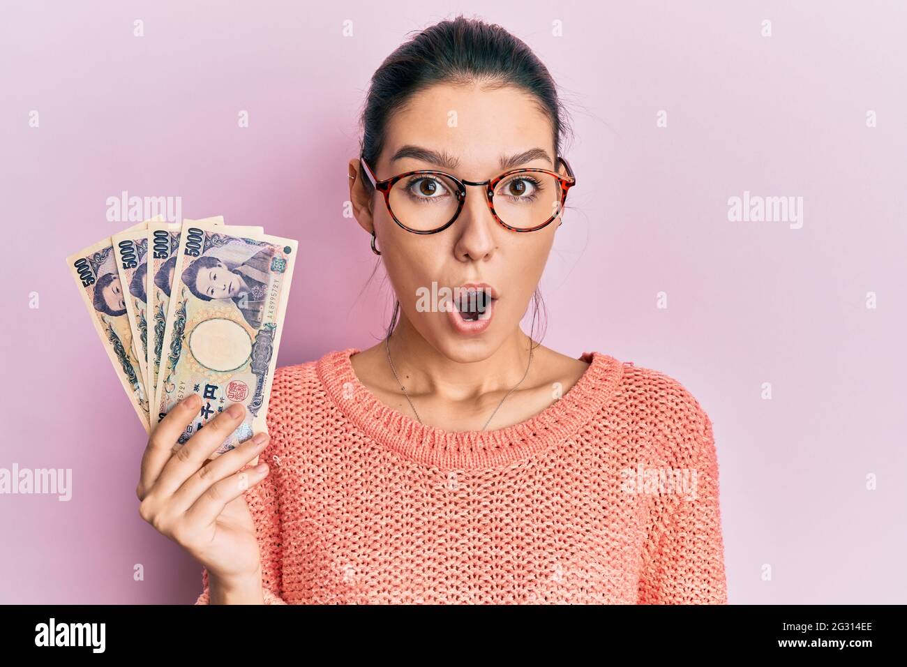 Young caucasian woman holding japanese yen banknotes scared and amazed ...