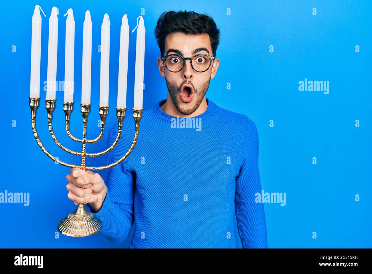 Young hispanic man holding menorah hanukkah jewish candle scared and ...