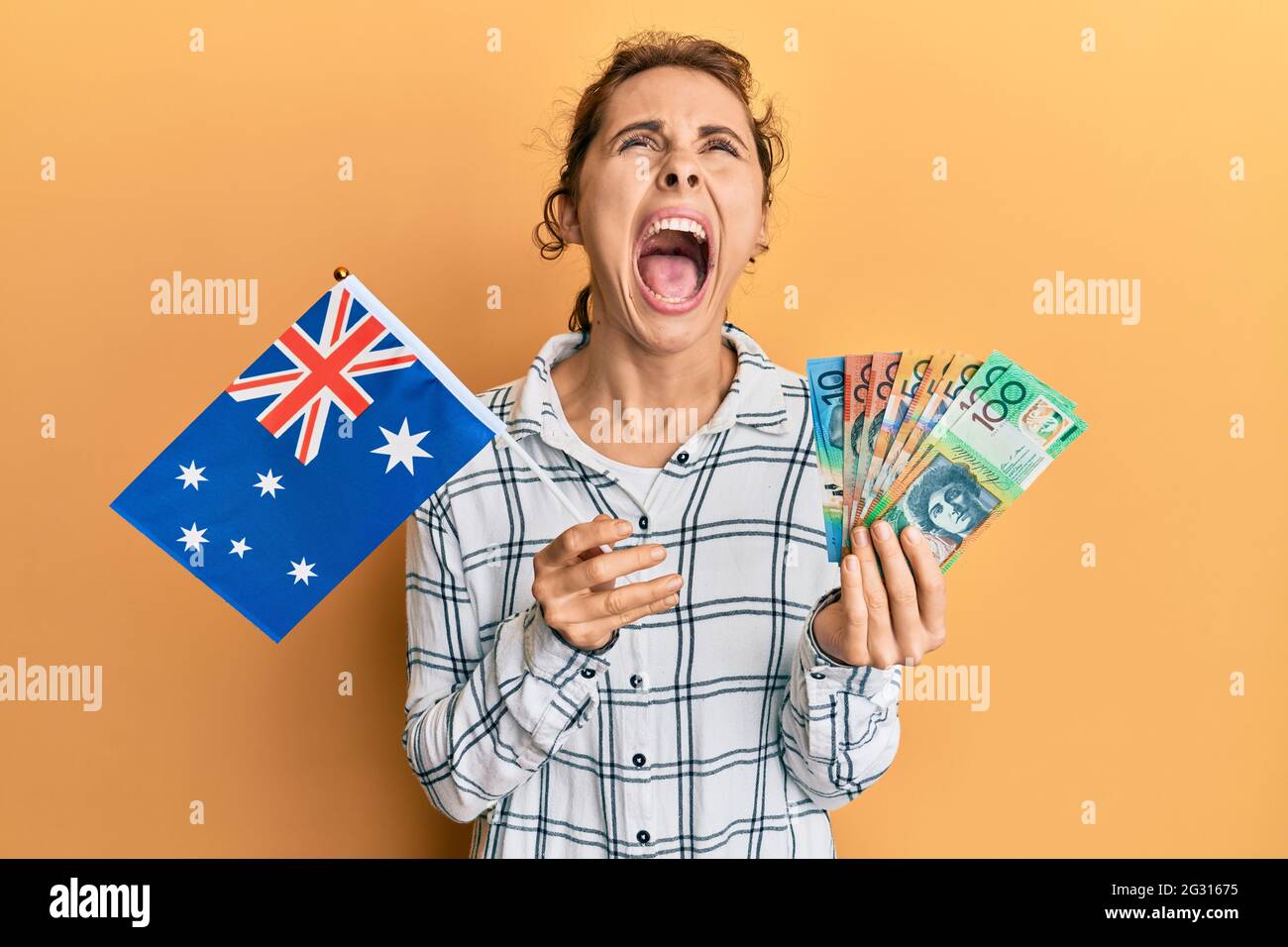 Young brunette woman holding australian flag and dollars angry and mad ...