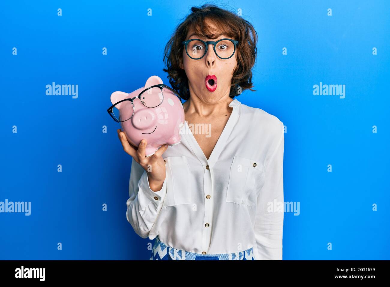 Young brunette woman holding piggy bank with glasses scared and amazed ...