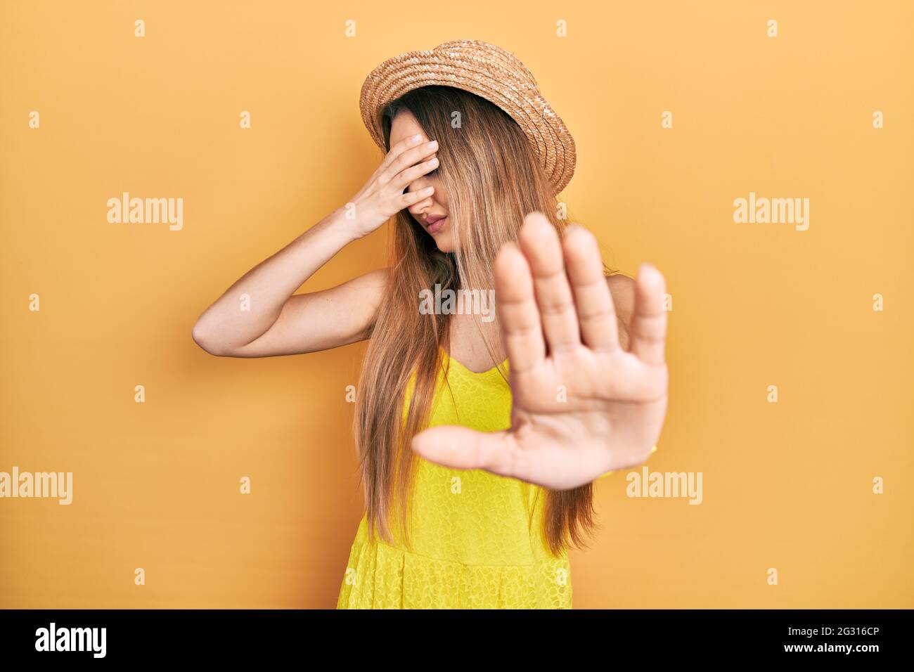 Young blonde girl wearing summer hat covering eyes with hands and doing ...