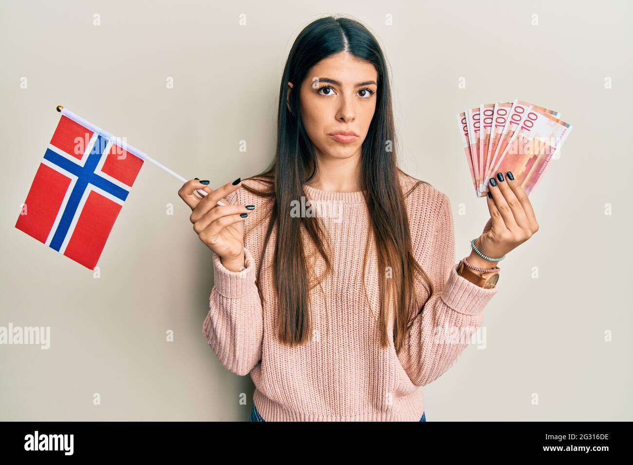 Young hispanic woman holding norway flag and krone banknotes depressed ...