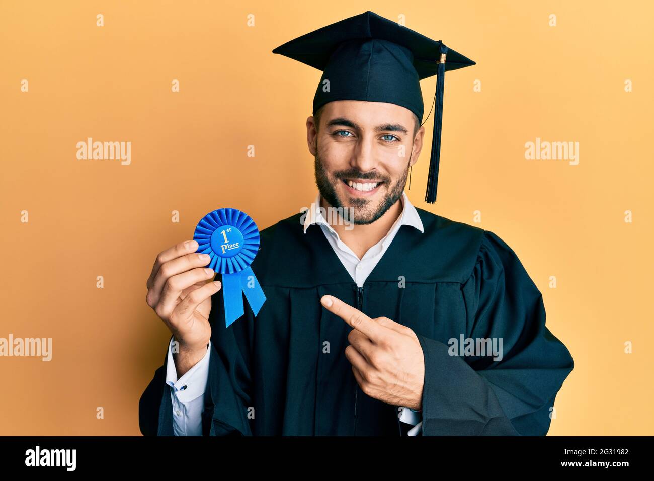 Young hispanic man wearing graduation robe holding 1 st place badge ...